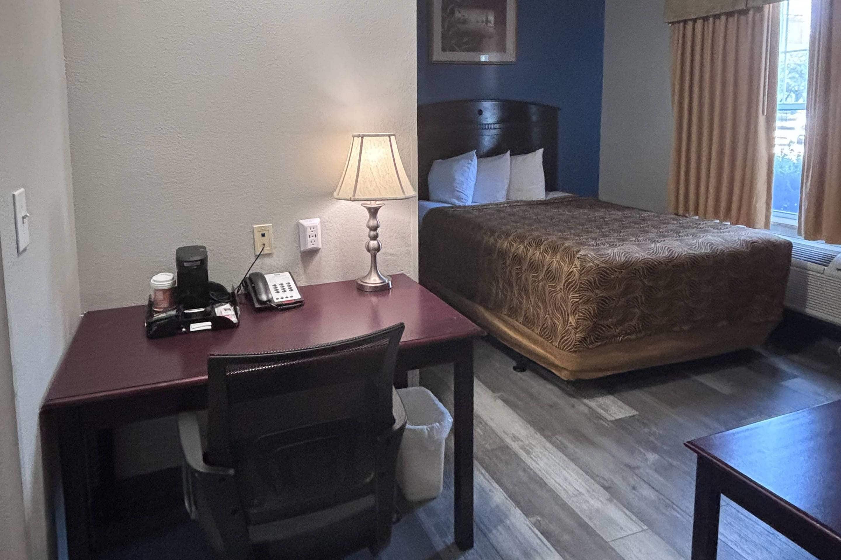 Quarto de hotel com cama, escrivaninha com luminária, cadeira, cafeteira e telefone.