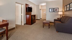Quarto de hotel com mesa de jantar de madeira, sofá, televisão de tela plana fixada em um móvel de madeira e pequena mesa lateral com abajur.