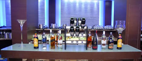 Uma área de bar com diversas garrafas de bebidas e copos, prateleiras com garrafas e uma grande garrafa de vidro para vinho.