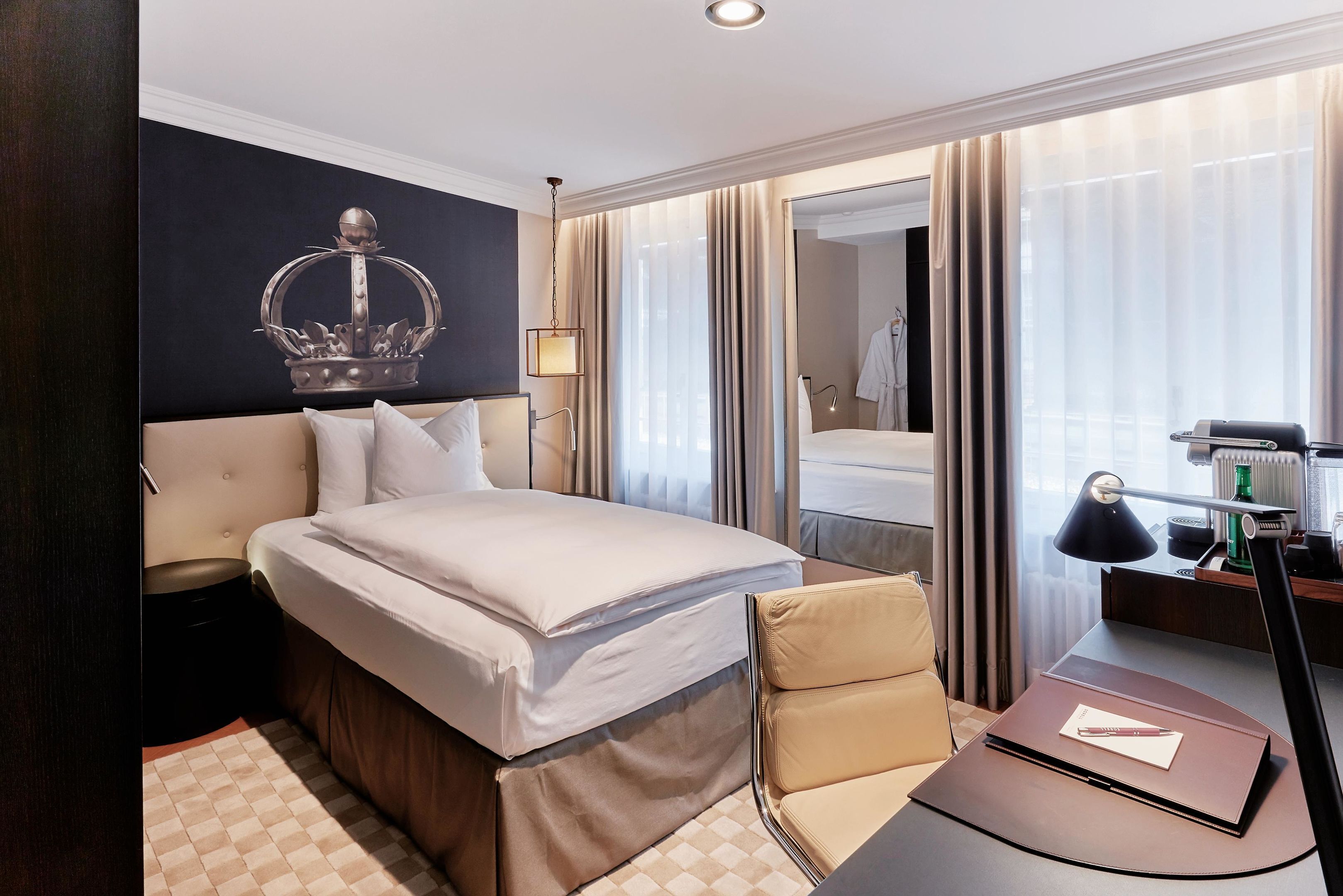 Quarto de hotel com uma cama, uma cadeira, uma escrivaninha, um espelho e uma janela com cortinas.