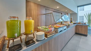Disposição de buffet com diversos alimentos e bebidas, incluindo um dispensador de líquido verde e um amarelo, além de um bar de saladas.