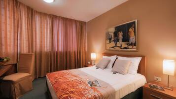 Quarto de hotel com uma cama grande, uma escrivaninha, uma cadeira, uma mesa de cabeceira e um quadro na parede.
