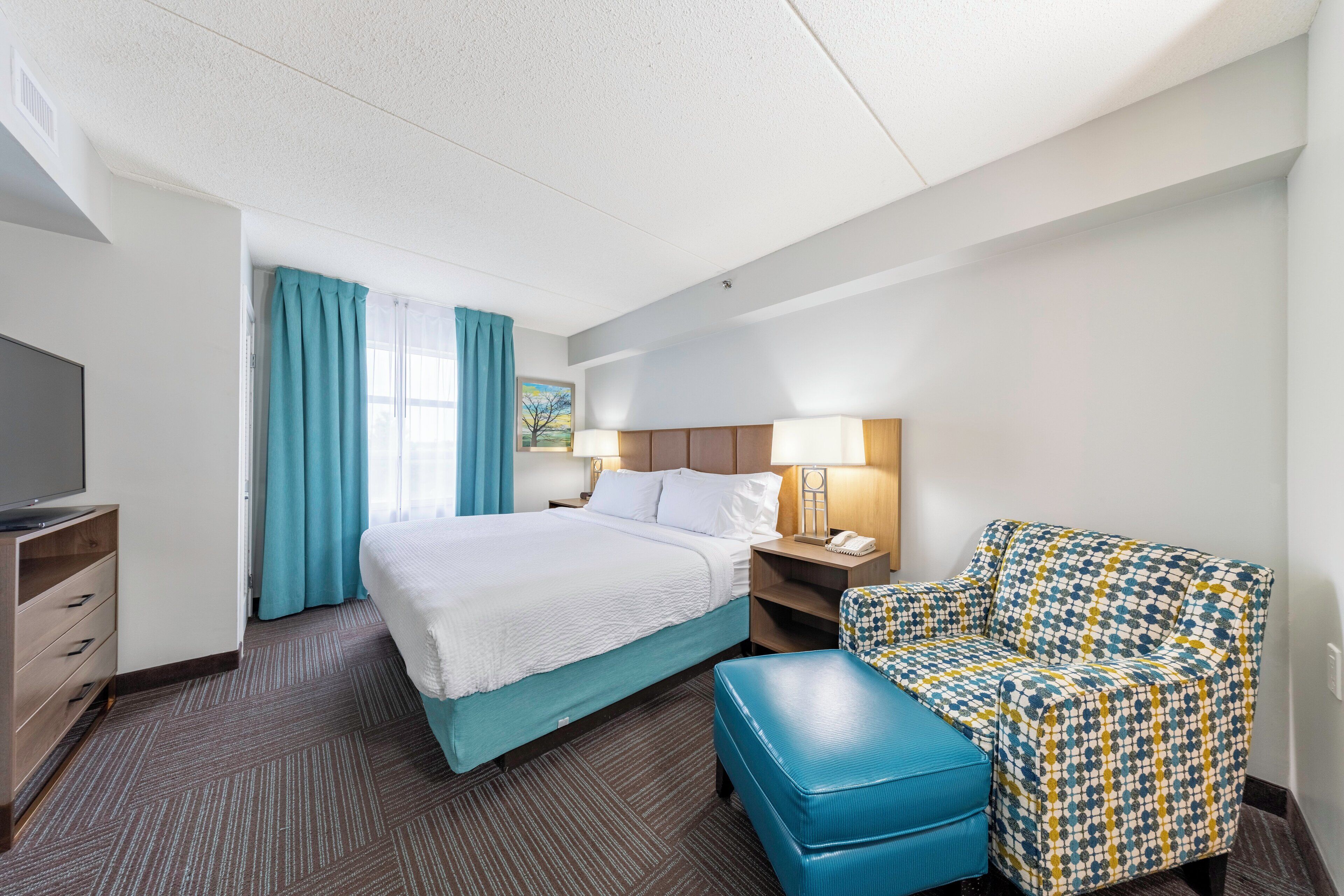 Quarto de hotel com cama, escrivaninha, cadeira, televisão e janela com cortinas.