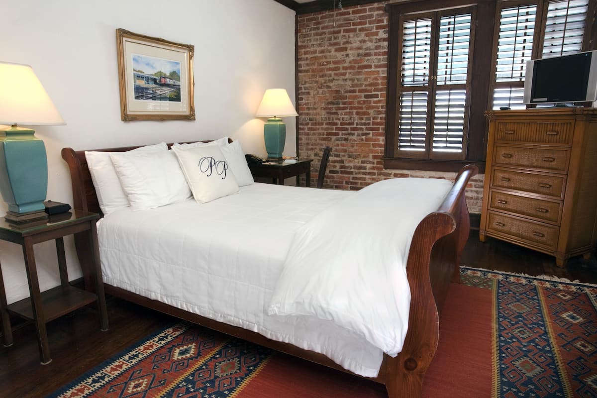 Uma cama bem arrumada com lençóis brancos e duas almofadas, um criado-mudo de madeira com uma luminária, um quadro na parede, uma parede de tijolos e um aparador de madeira com uma televisão.