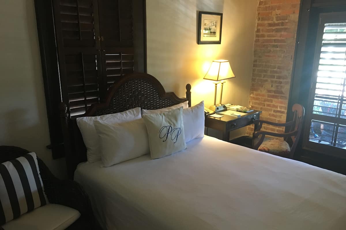 Quarto de hotel com cama, escrivaninha com luminária, cadeira e janela com persianas.