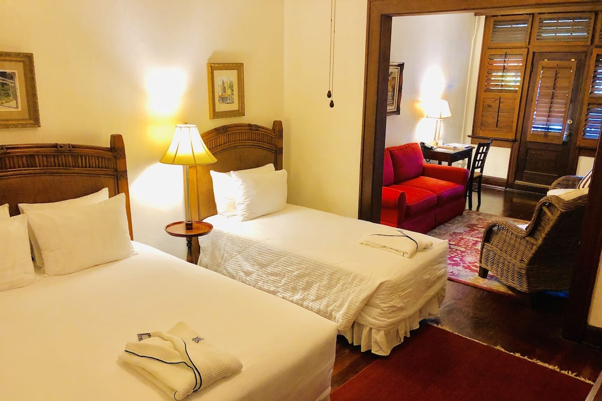 Quarto de hotel com duas camas, uma poltrona vermelha, uma cadeira de vime e uma porta de madeira com portaria.