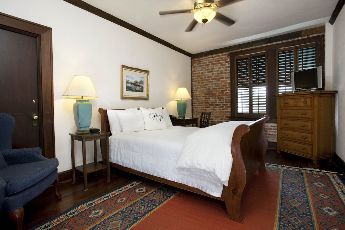 Quarto com cama de madeira, duas luminárias de cabeceira, cômoda de madeira, poltrona azul, tapete estampado e ventilador de teto.