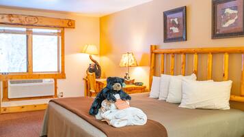 Quarto de hotel com cama, um urso de pelúcia, abajur e janela.
