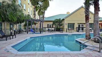 Piscina com espreguiçadeiras e um pérgola, cercada por palmeiras e um edifício com telhado verde.