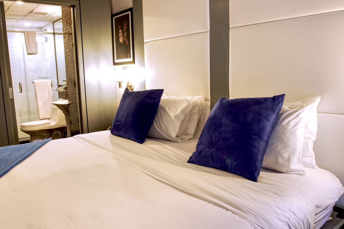 Quarto de hotel com cama, duas almofadas, mesa de cabeceira, espelho e banheiro visível por uma porta aberta.