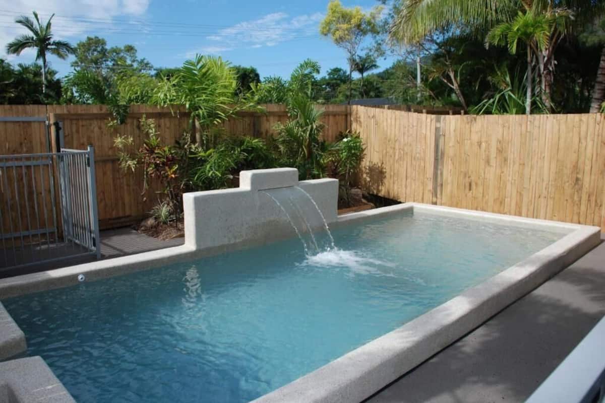 Uma piscina retangular com uma cascata, cercada por um muro de madeira e plantas tropicais.