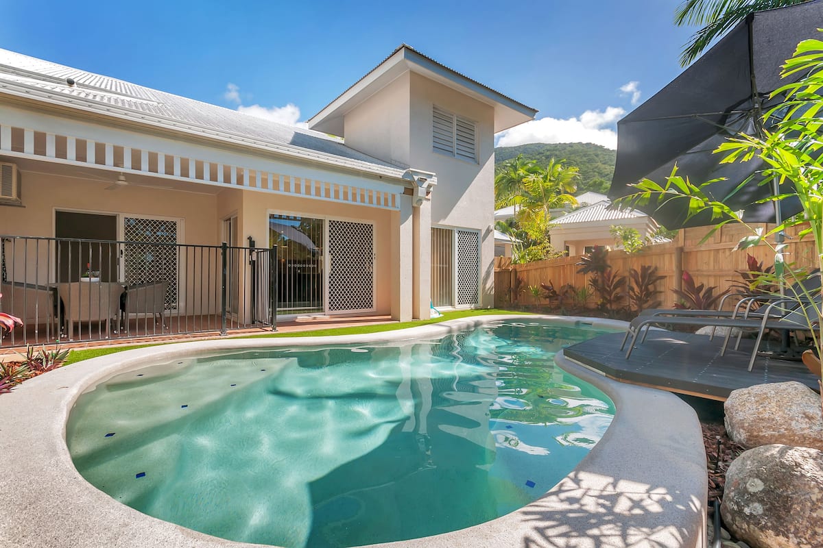 Piscina com hidromassagem, cercada por uma casa com varanda e cerca de madeira.