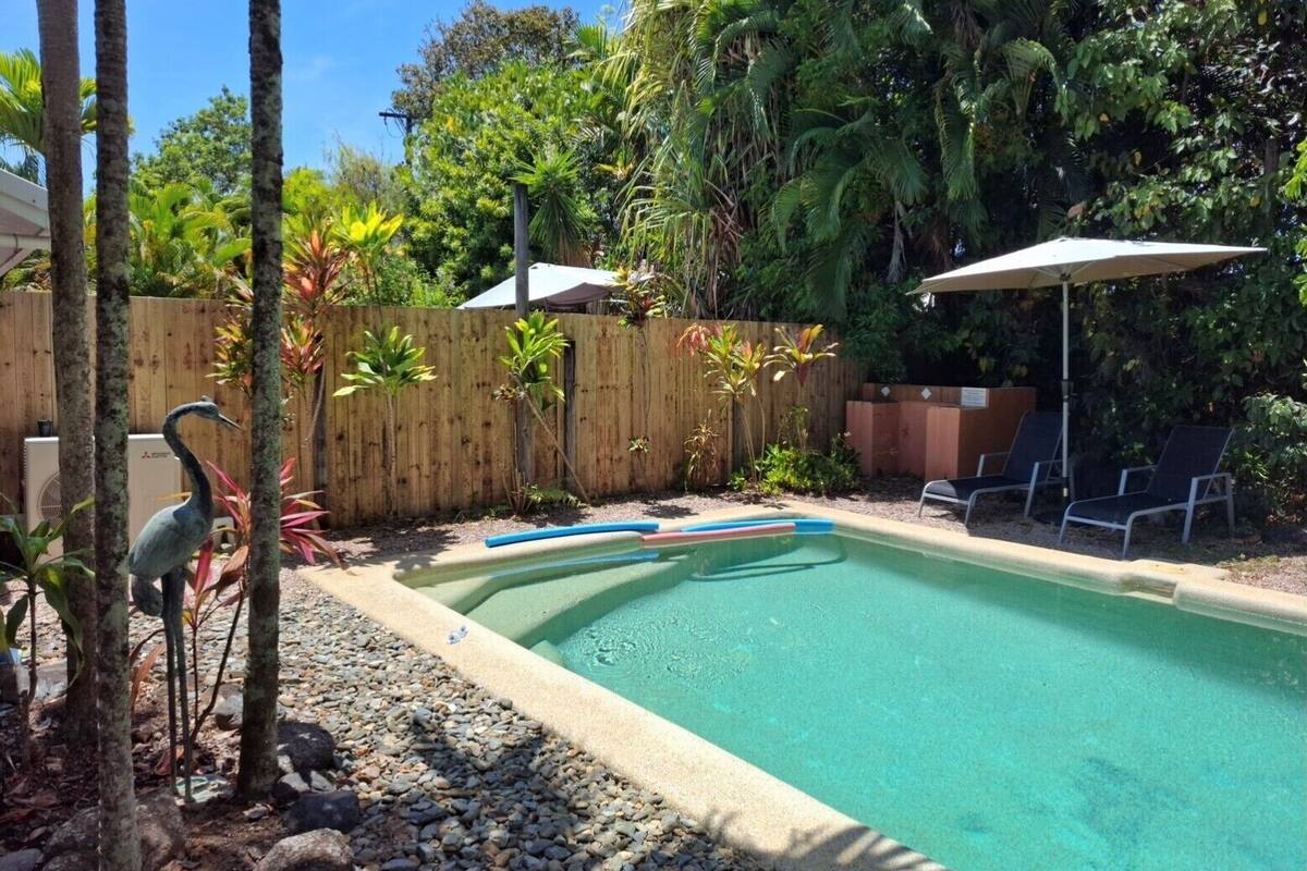 Um quintal com piscina, cercado por um muro de madeira, palmeiras e uma estátua de pássaro.