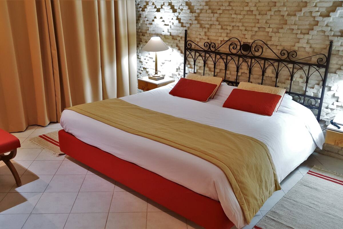 Uma cama bem arrumada com roupa de cama vermelha e bege, uma mesinha de cabeceira de madeira com abajur, e uma parede de tijolos ao fundo.