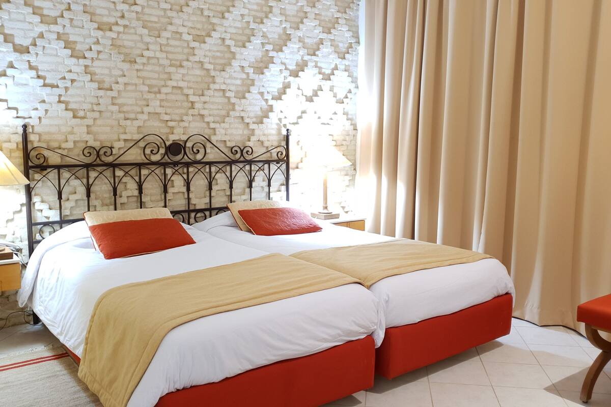 Uma cama de casal com roupa de cama vermelha e branca, um cobertor bege e um travesseiro vermelho. Há uma mesa de cabeceira com uma luminária e uma pequena mesa com uma vela. O quarto tem uma parede de tijolos e uma janela com cortinas.