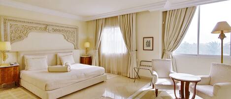 Quarto de hotel com uma cama grande, área de estar com uma cadeira e mesa, mesa de cabeceira com abajur e janela com cortinas.