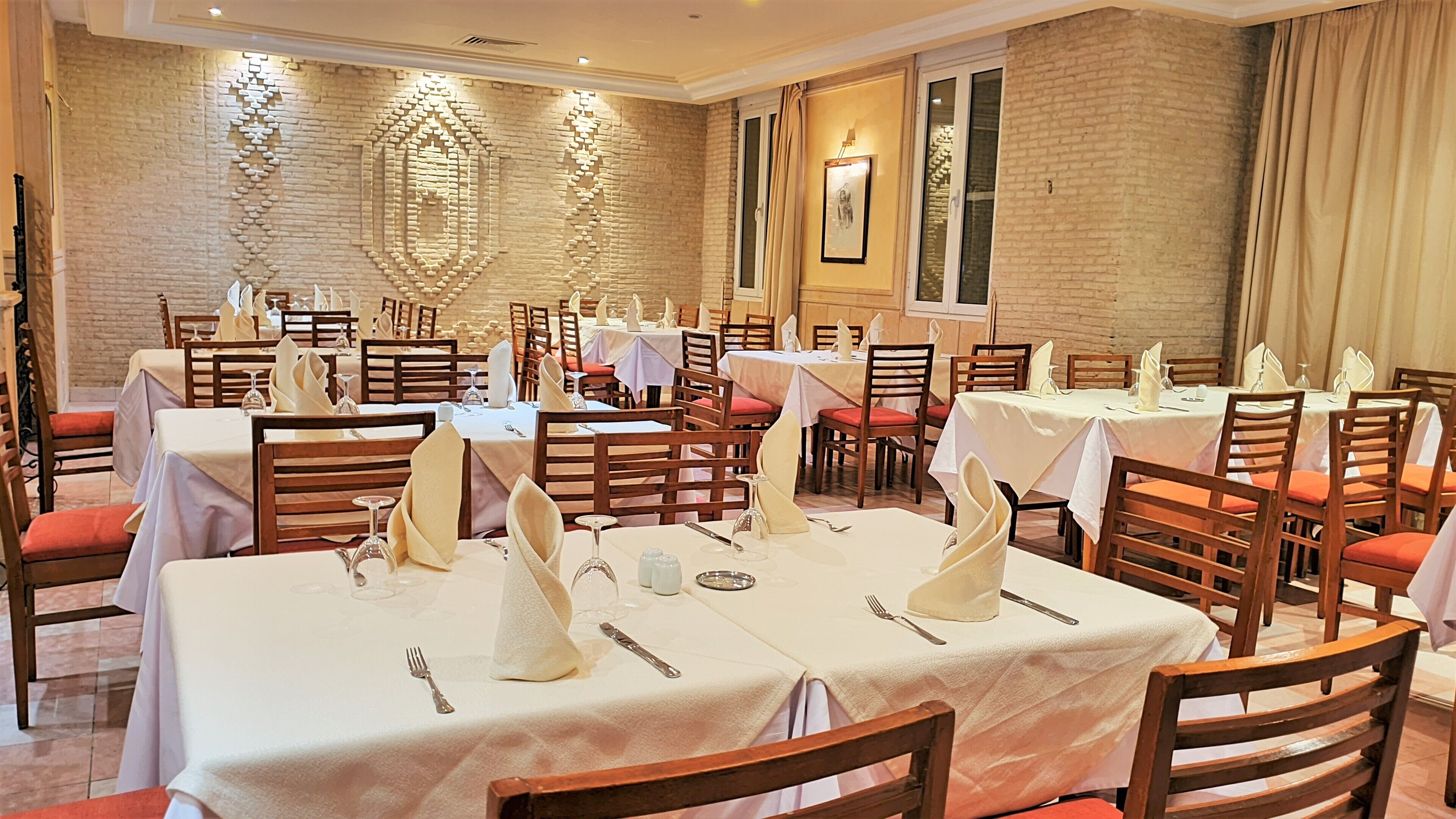 Um restaurante com várias mesas e cadeiras, toalhas de mesa brancas e uma parede de tijolos com iluminação decorativa.