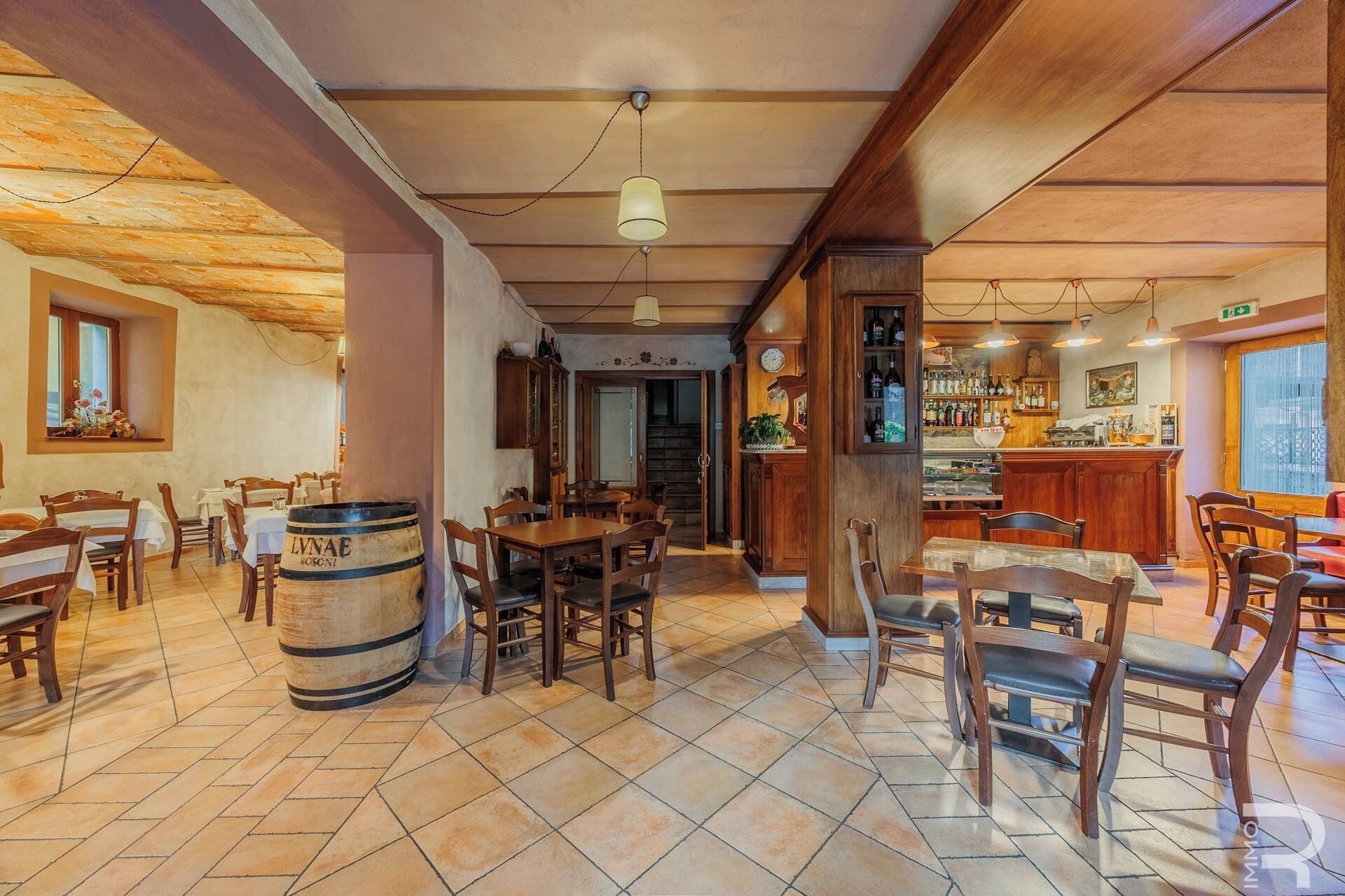 Restaurante com mesas e cadeiras de madeira, um espaço de bar e piso ladrilhado.