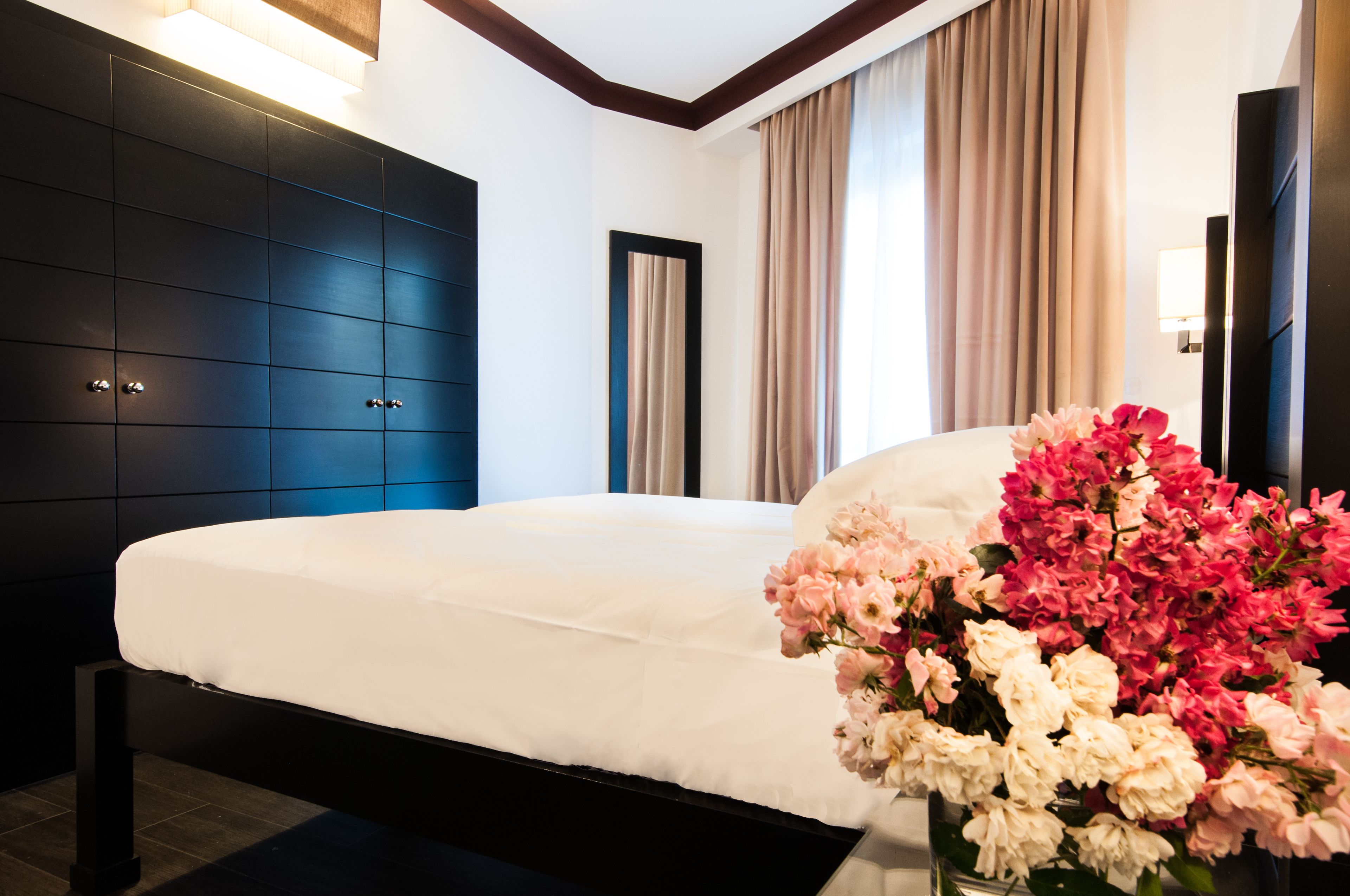 Quarto de hotel com uma cama grande, guarda-roupa azul escuro, cortinas bege e um vaso de flores.