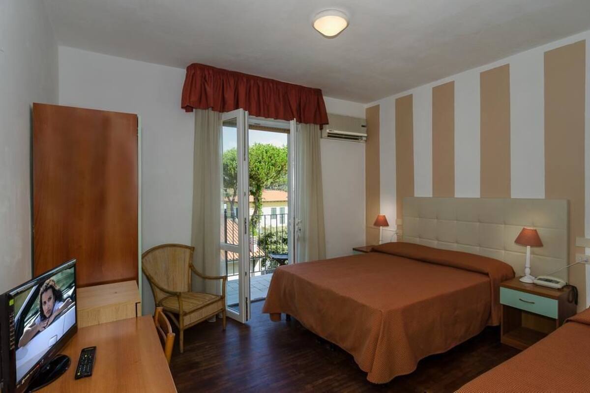 Quarto de hotel com cama, cadeira, escrivaninha com TV e varanda com vista.