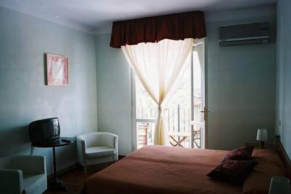Quarto de hotel com cama, cadeira, televisão e janela com cortinas.