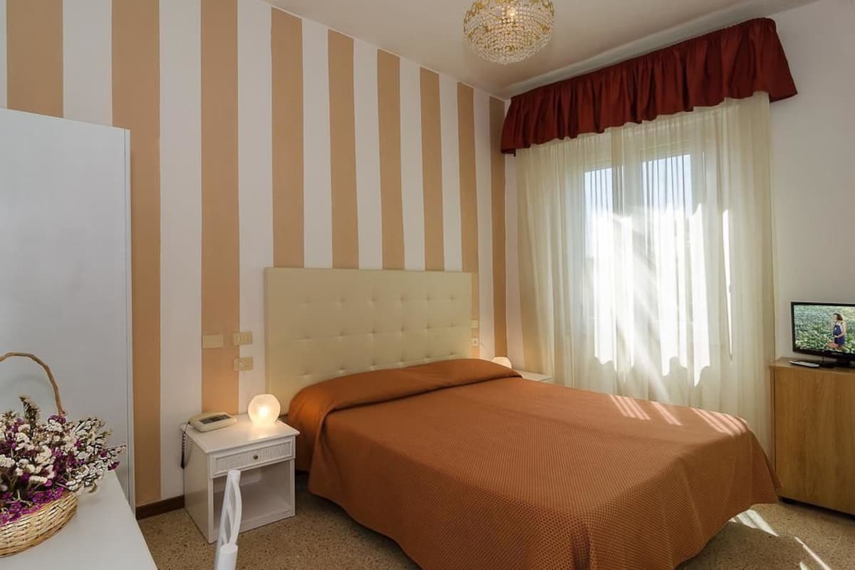 Quarto de hotel com parede listrada, uma cama com colcha marrom, mesa de cabeceira com abajur, televisão em móvel de madeira e janela com cortinas leves.