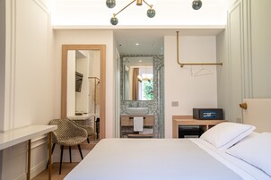 Quarto de hotel moderno com uma cama grande, uma mesa com cadeira, toucador com espelho e banheiro com pia e chuveiro.