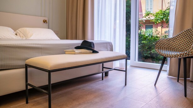 Quarto moderno com cama, um banco e uma cadeira, grandes janelas com cortinas, e vista para edifícios lá fora.