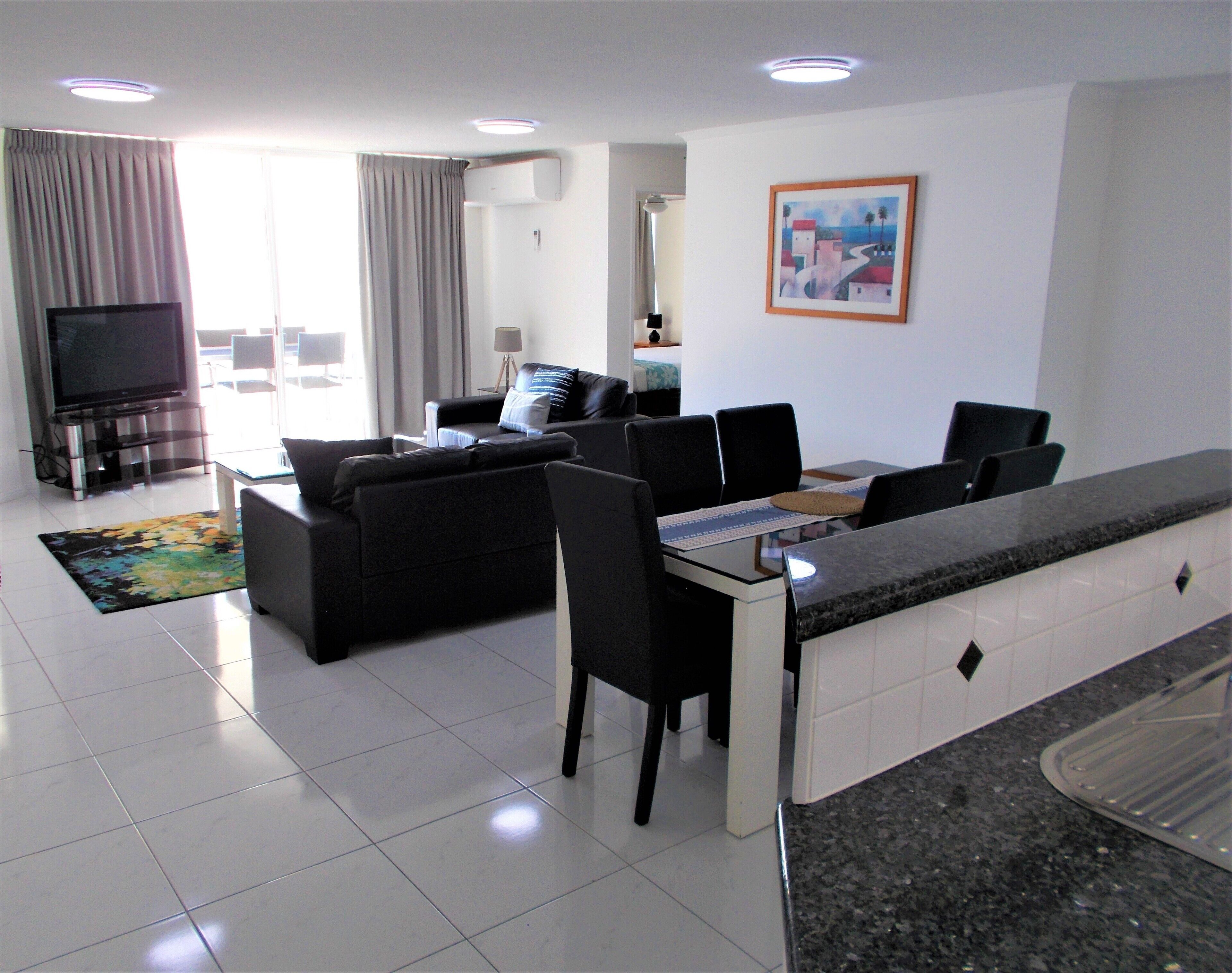 Uma sala de estar moderna com televisão de tela plana, um sofá, área de refeições com mesa e cadeiras, e cozinha com bancada de granito.