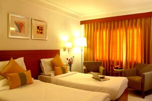 Quarto de hotel com duas camas, uma escrivaninha com um laptop, uma cadeira e uma mesa com uma tigela de frutas.