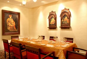 Uma sala de jantar com uma mesa comprida posta para seis pessoas, rodeada por cadeiras estofadas vermelhas. Há duas pinturas decorativas penduradas na parede acima da mesa.