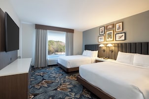 Quarto de hotel com duas camas, uma escrivaninha e uma janela grande com cortinas.
