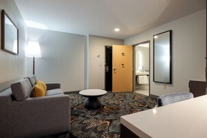 Quarto de hotel com sofá cinzento, mesa redonda, tapete estampado e espelho na parede.