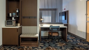 Quarto de hotel pequeno com cozinha compacta, escrivaninha, cadeira, janela grande com persianas e carpete estampado.