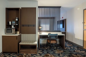 Quarto de hotel pequeno com cozinha compacta, escrivaninha, cadeira, janela grande com persianas e carpete estampado.