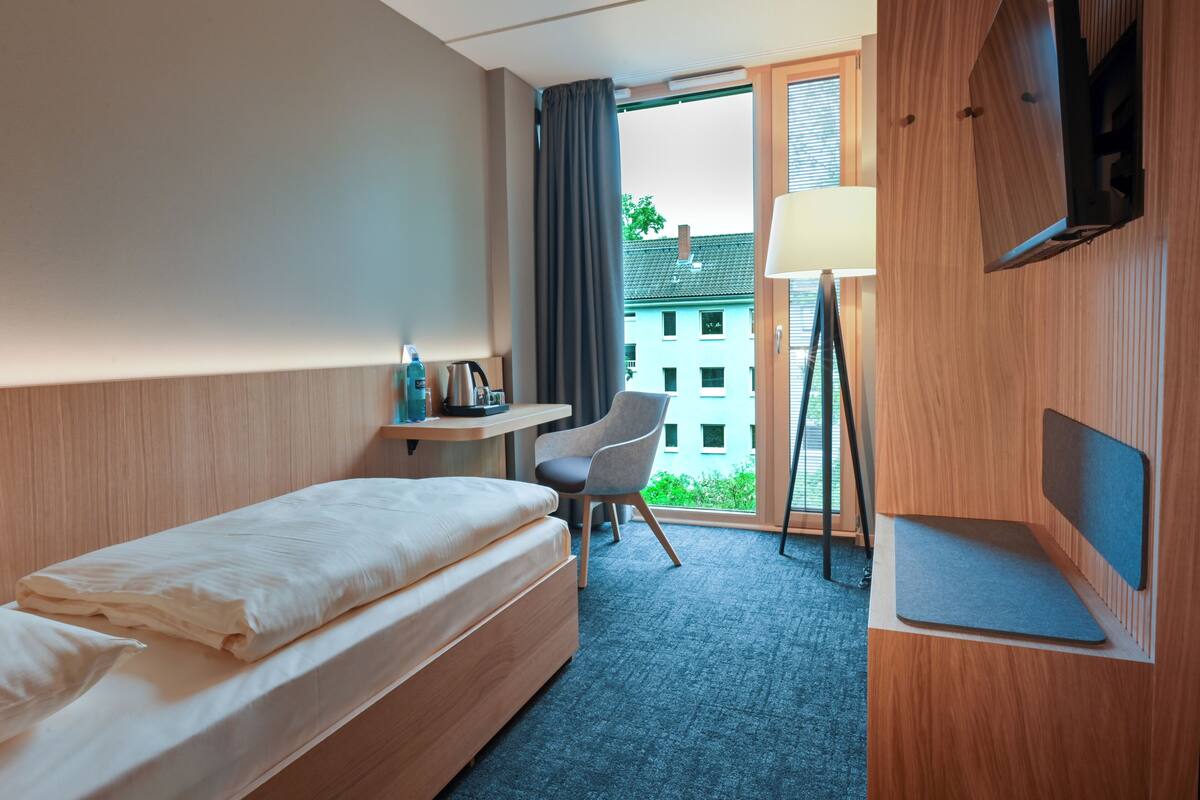 Quarto de hotel com cama, escrivaninha, cadeira, abajur e televisão fixada na parede.