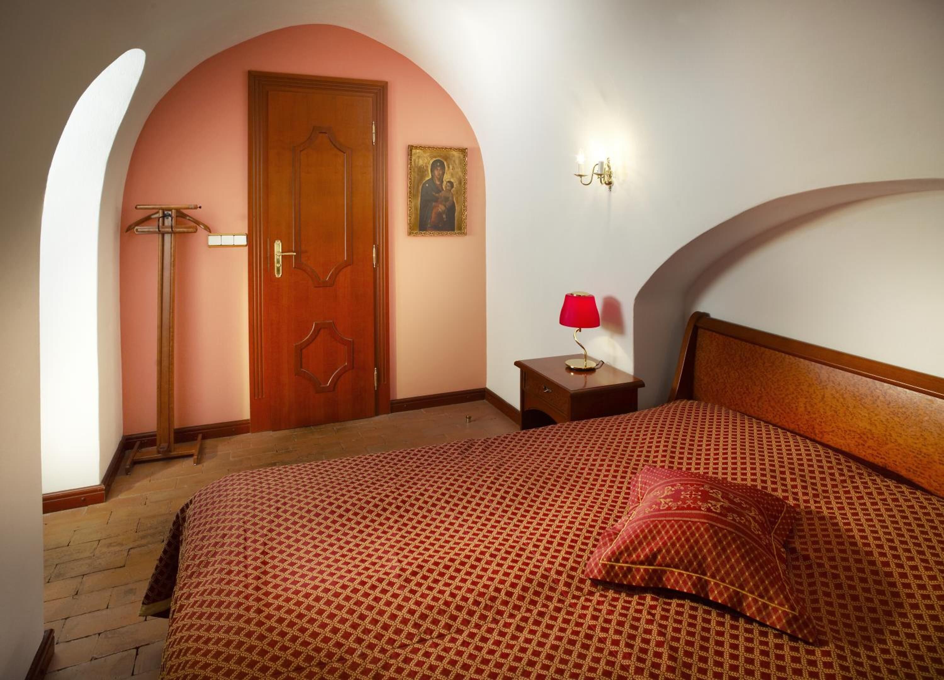 Quarto com colcha estampada, porta de madeira, luminária na parede e pequena mesa de cabeceira.