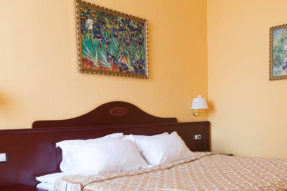Quarto de hotel com uma cama, duas pinturas nas paredes, uma mesa de cabeceira e um abajur.