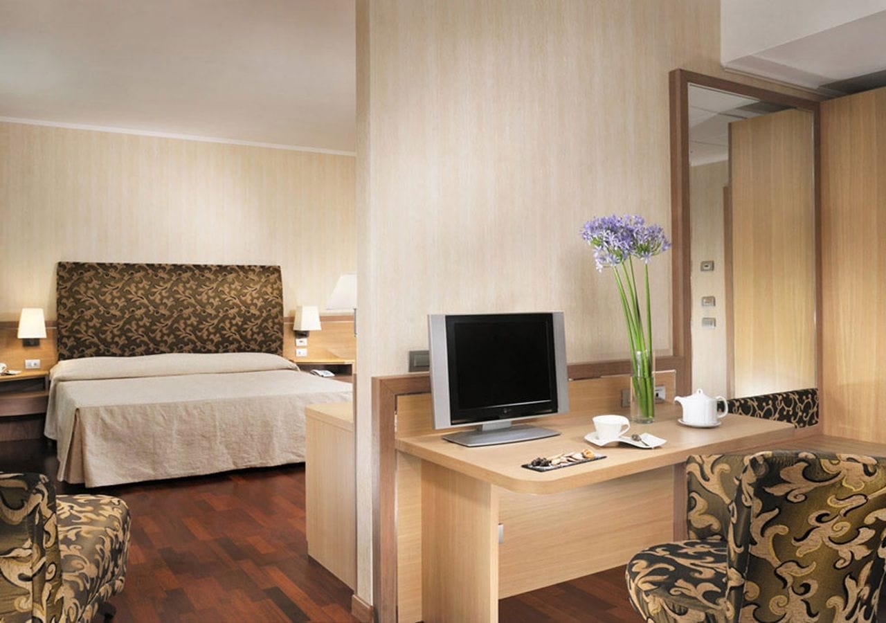 Quarto de hotel com uma cama, uma escrivaninha com computador, uma cadeira e um espelho.