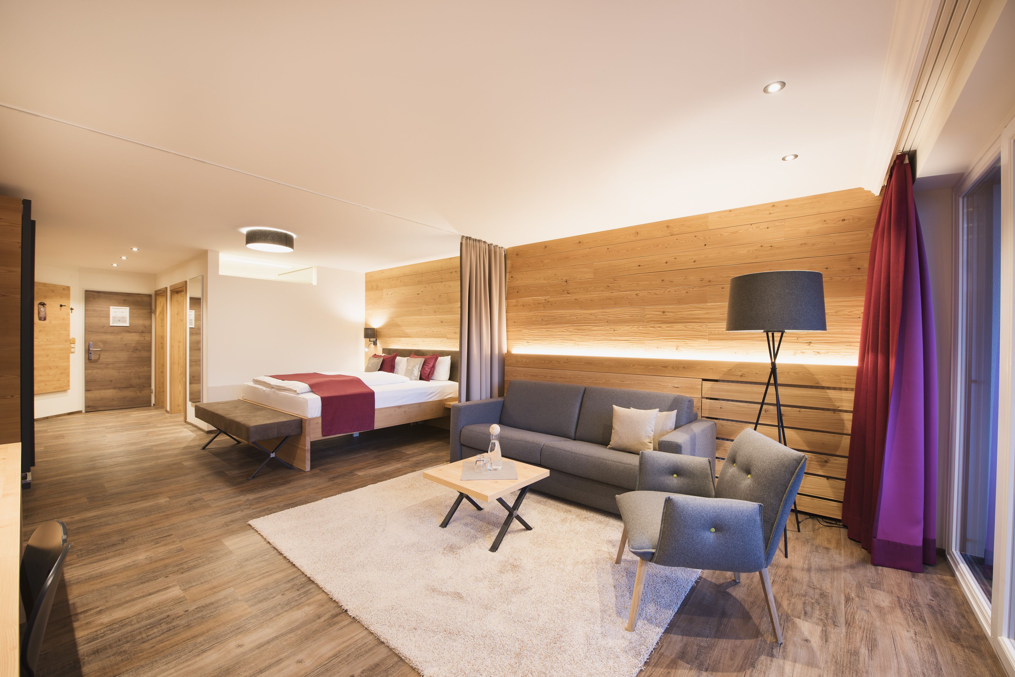 Quarto de hotel moderno com cama, sofá, poltrona e uma luminária.