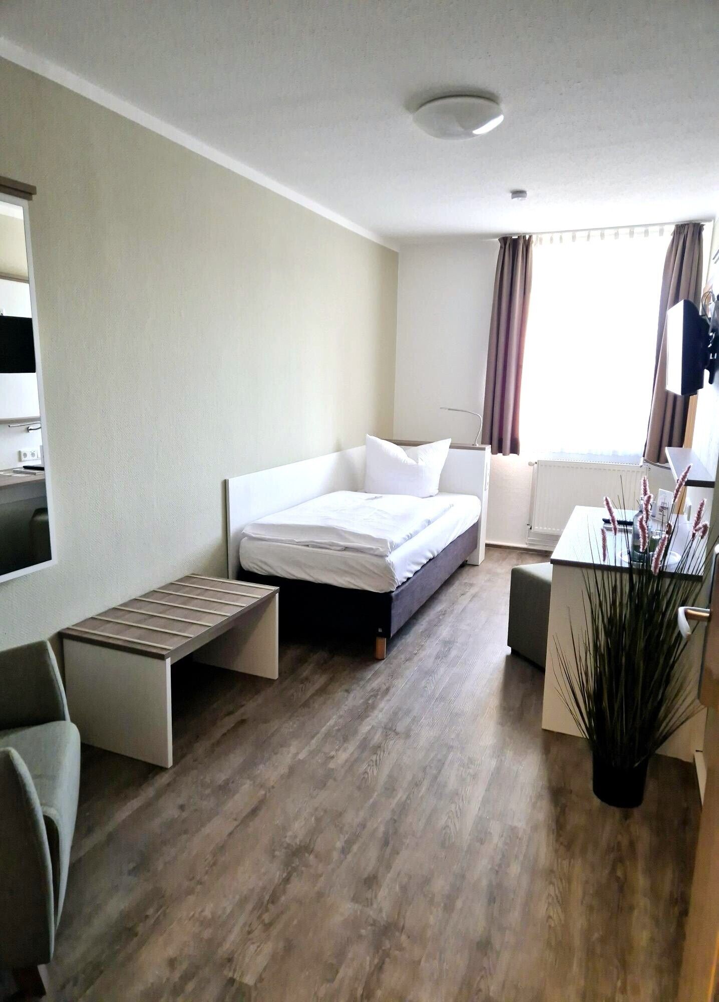 Quarto de hotel moderno com cama, escrivaninha, cadeira, mesinha e planta em vaso.