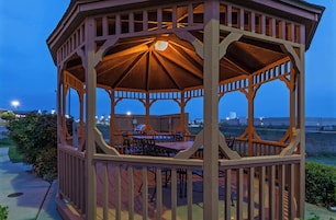 Um gazebo de madeira com telhado, rodeado por um deck com cadeiras e mesas, com vista para um corpo d'água.