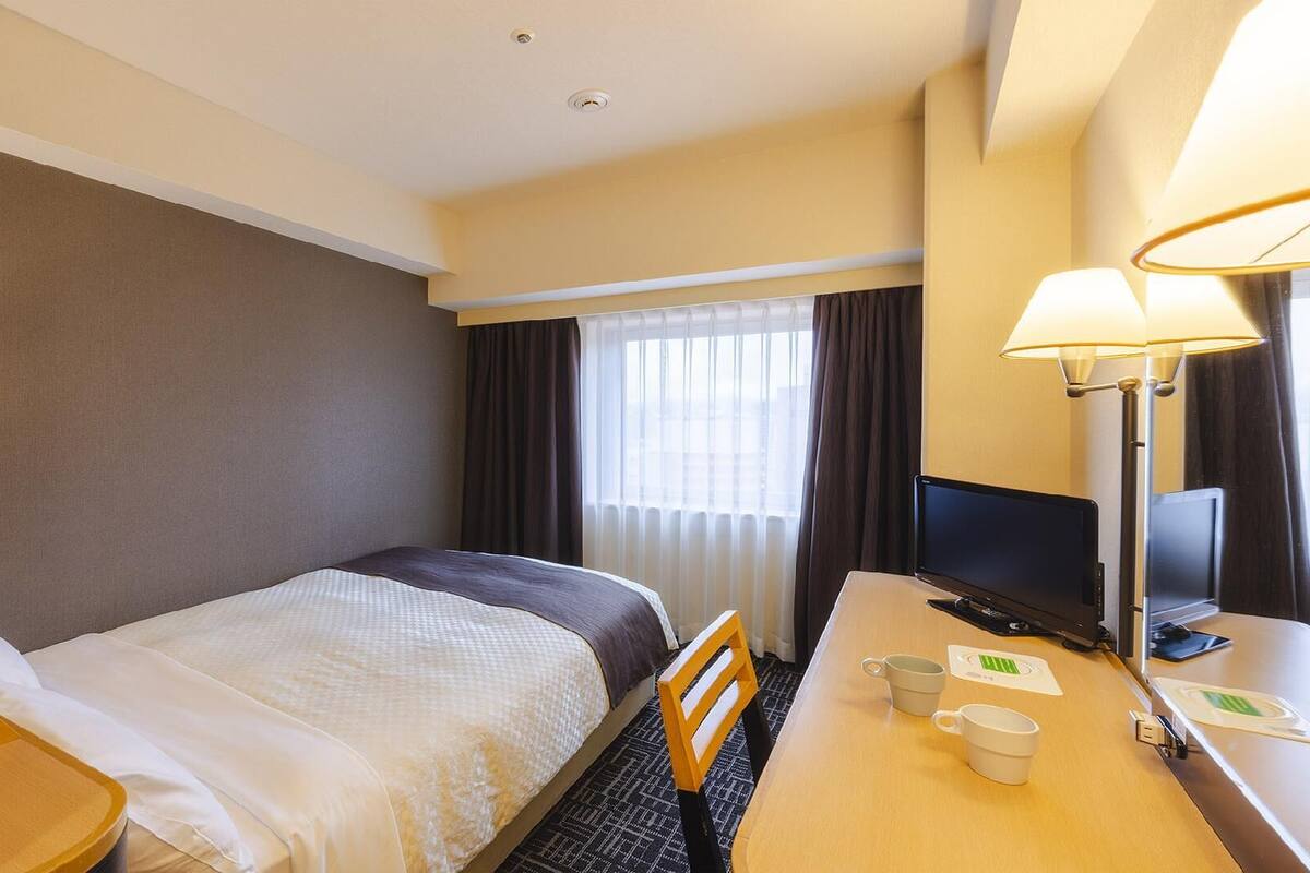 Quarto de hotel com uma cama, uma escrivaninha com computador, uma cadeira e uma luminária.