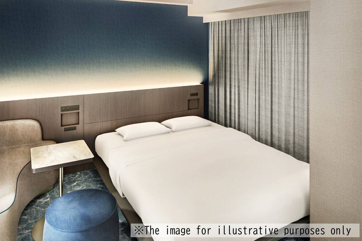 Quarto de hotel com cama, uma pequena mesa, uma cadeira e um banquinho azul.