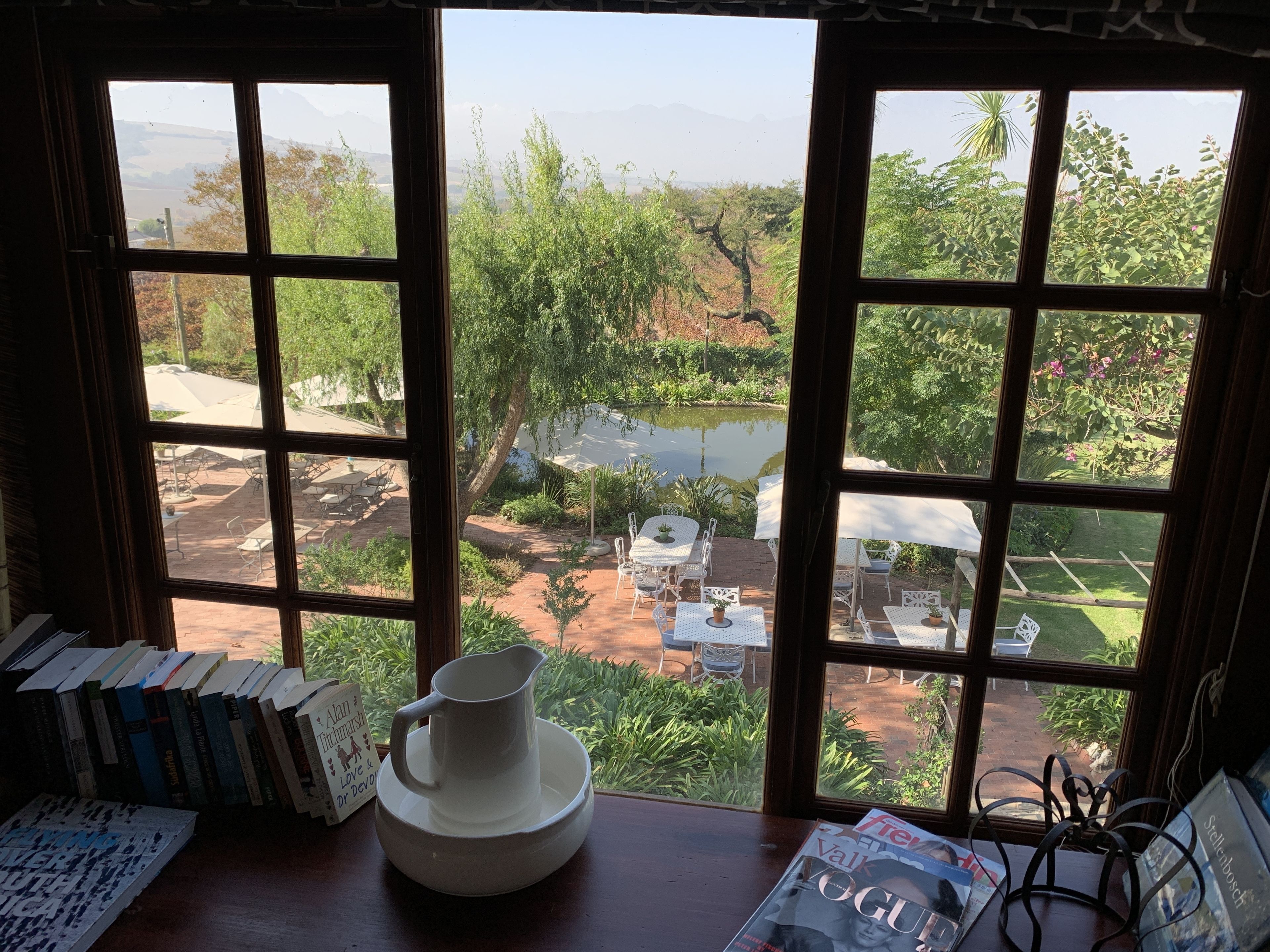 Visão pela janela, mostrando uma mesa com livros e um jarro branco.