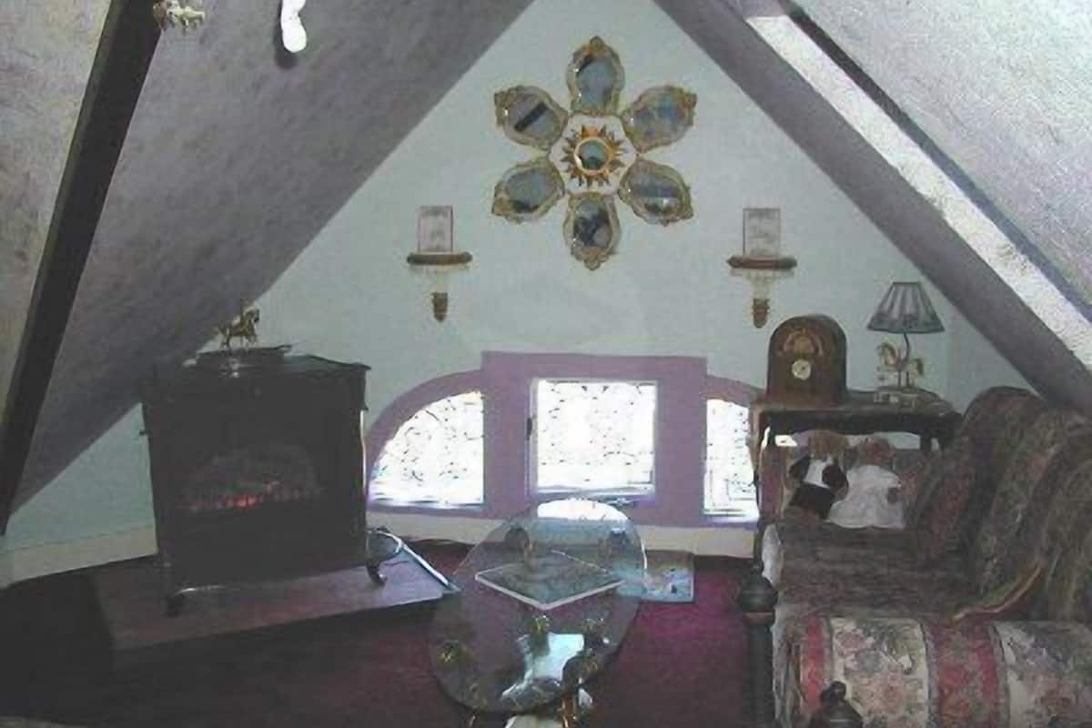 Um quarto de sótão acolhedor, com teto inclinado, uma moldura decorativa no teto, um armário de madeira, uma mesa de vidro, um sofá estampado e uma janela com cortinas.