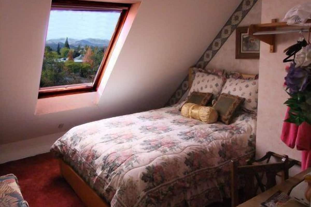 Quarto com teto inclinado, cama com roupa de cama floral, janela com vista para árvores e montanhas, e uma cadeira de madeira.