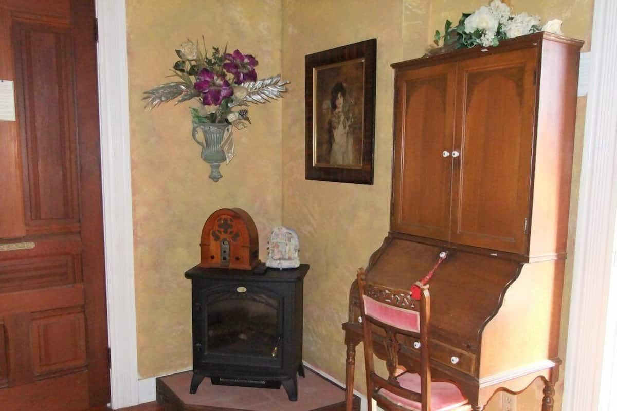 Um quarto com porta de madeira, um pequeno armário de madeira, uma cadeira de madeira com almofada, uma prateleira fixa com flores e um quadro emoldurado.