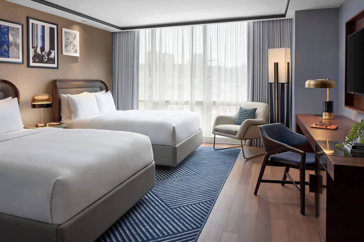 Quarto de hotel com duas camas, uma escrivaninha com cadeira, televisão e obras de arte na parede.