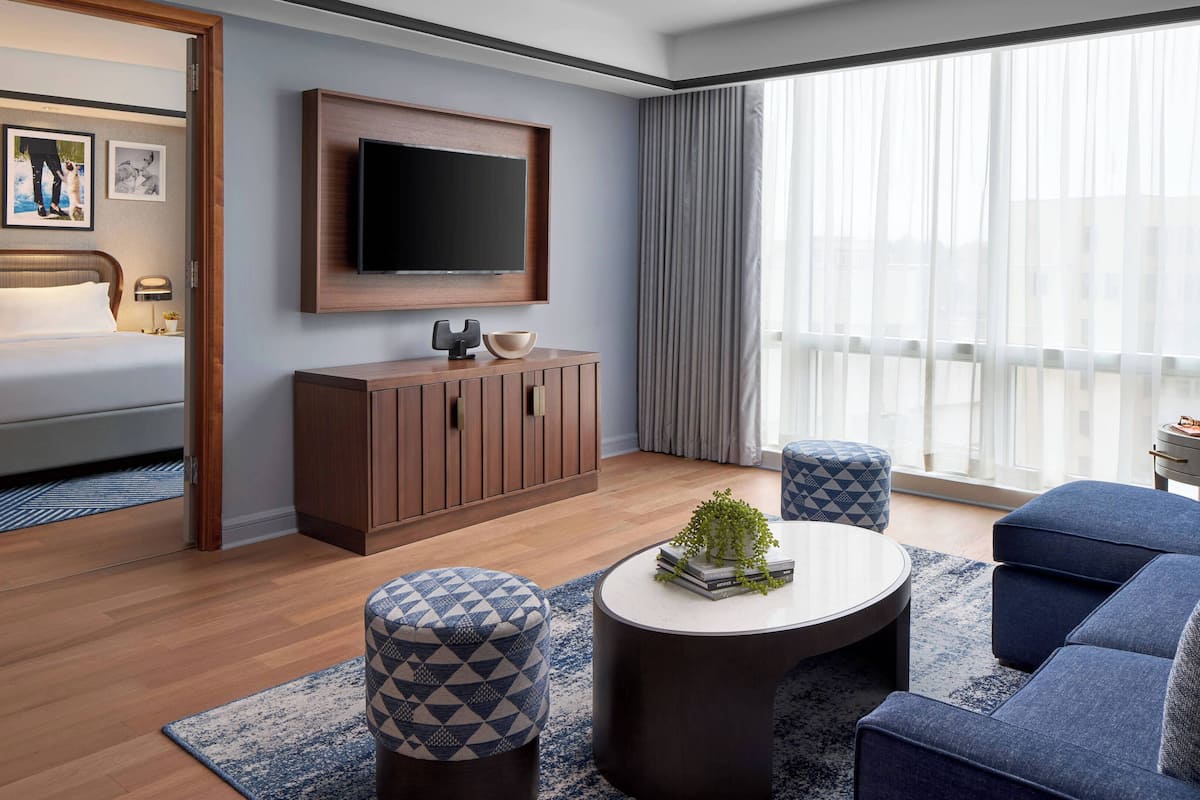 Quarto de hotel moderno com cama, televisão fixada na parede, área de estar com sofás e pufes, e uma janela grande com cortinas.